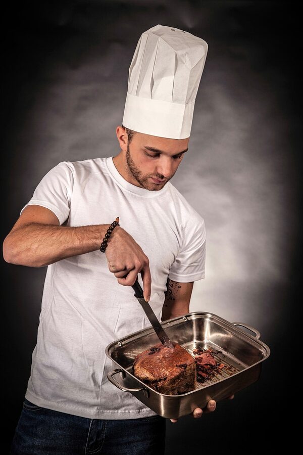 Chef in hat cooking and cutting meat with a pan in kitchen