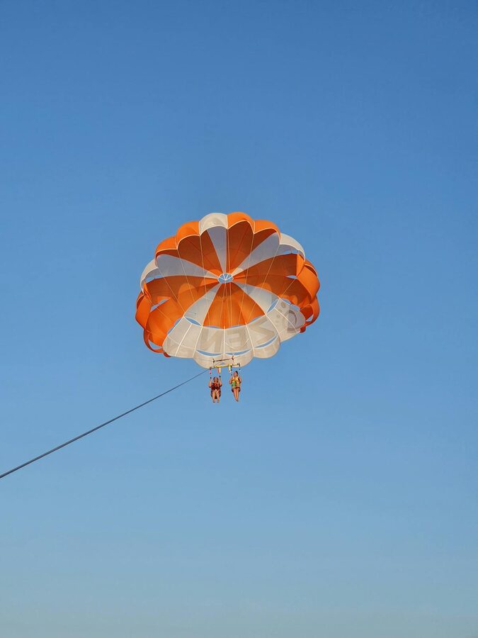People parasailing under a bright orange parachute against clear blue sky