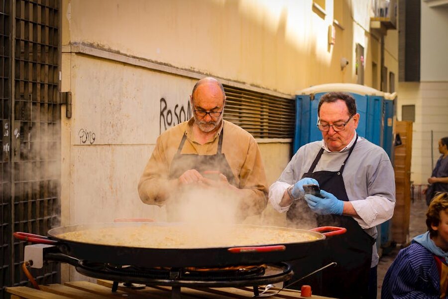Two chefs cooking a large paella outdoors with a phone recipe